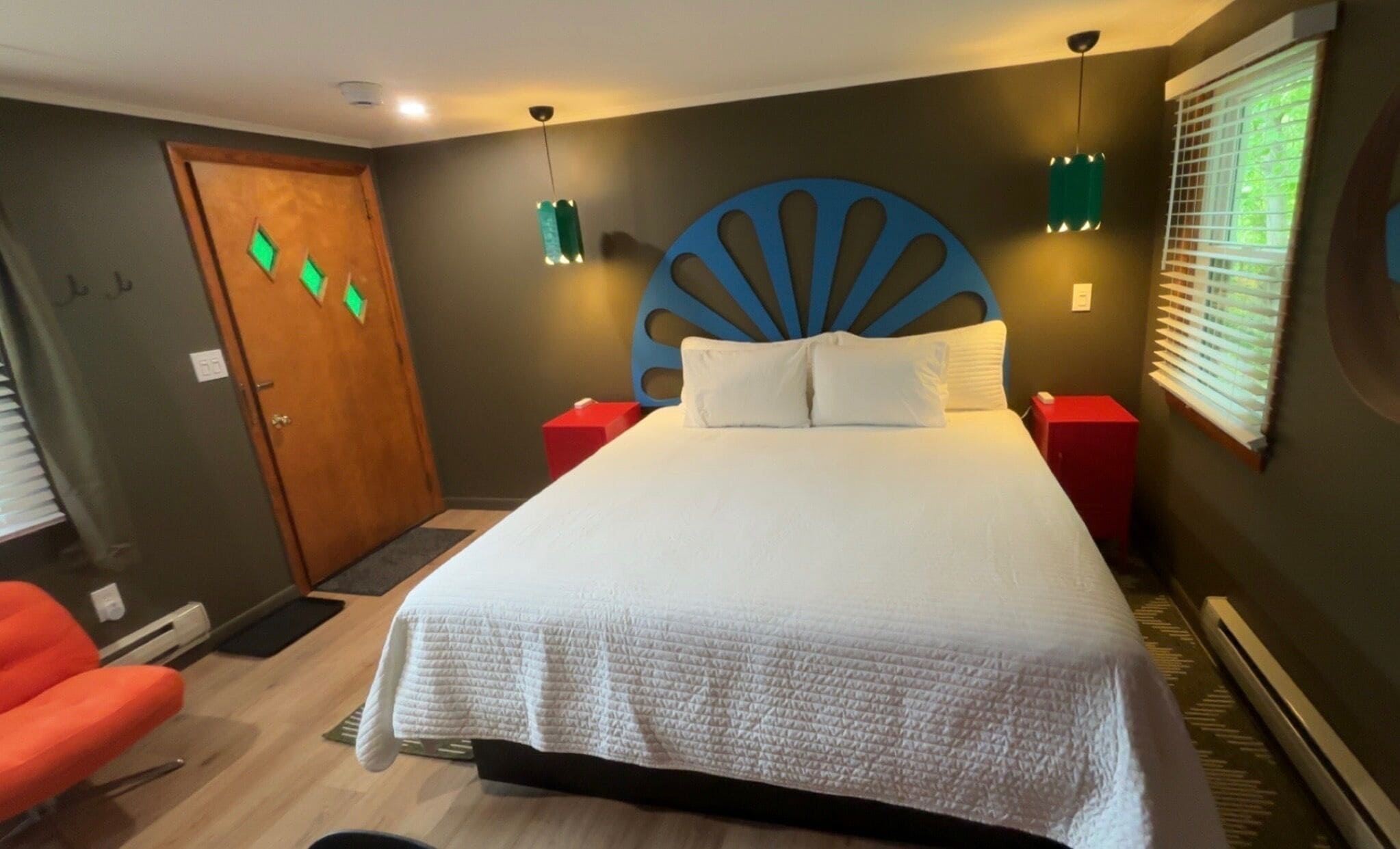 Um quarto com cama, porta, uma cadeira vermelha e janela com persianas.