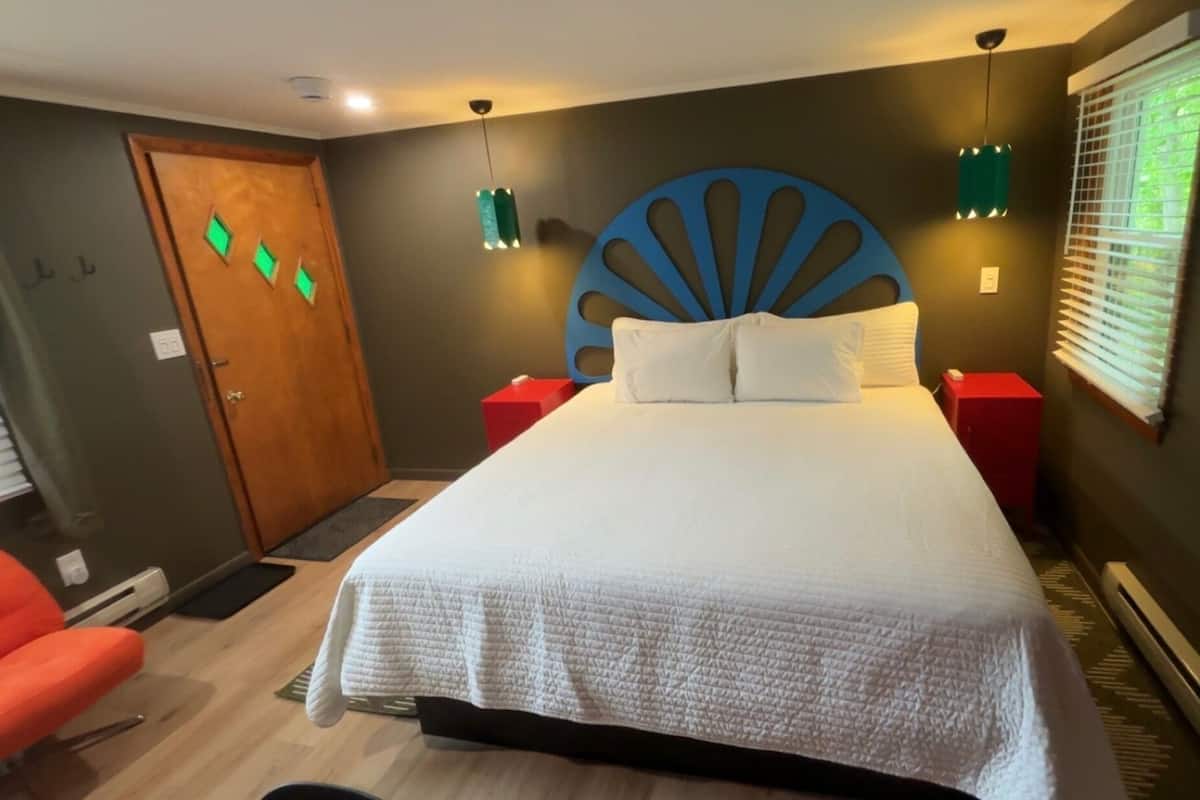 Um quarto com cama, porta, uma cadeira vermelha e janela com persianas.