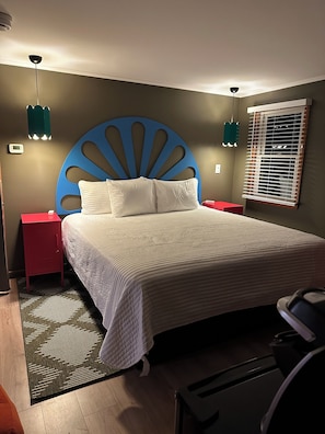 Uma cama com uma cabeceira azul distinta, uma mesa de cabeceira vermelha, um tapete verde e uma janela com persianas.