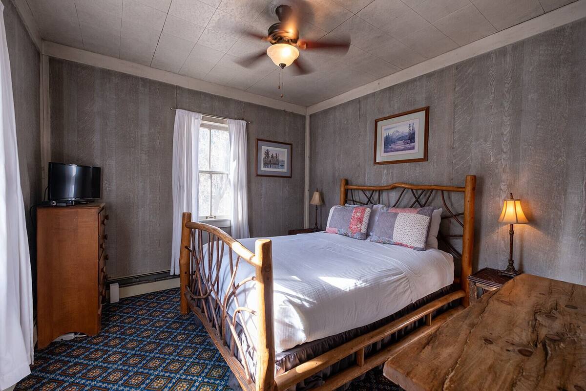 Quarto com cama de madeira, ventoinha no teto, televisão sobre um aparador e chão estampado.