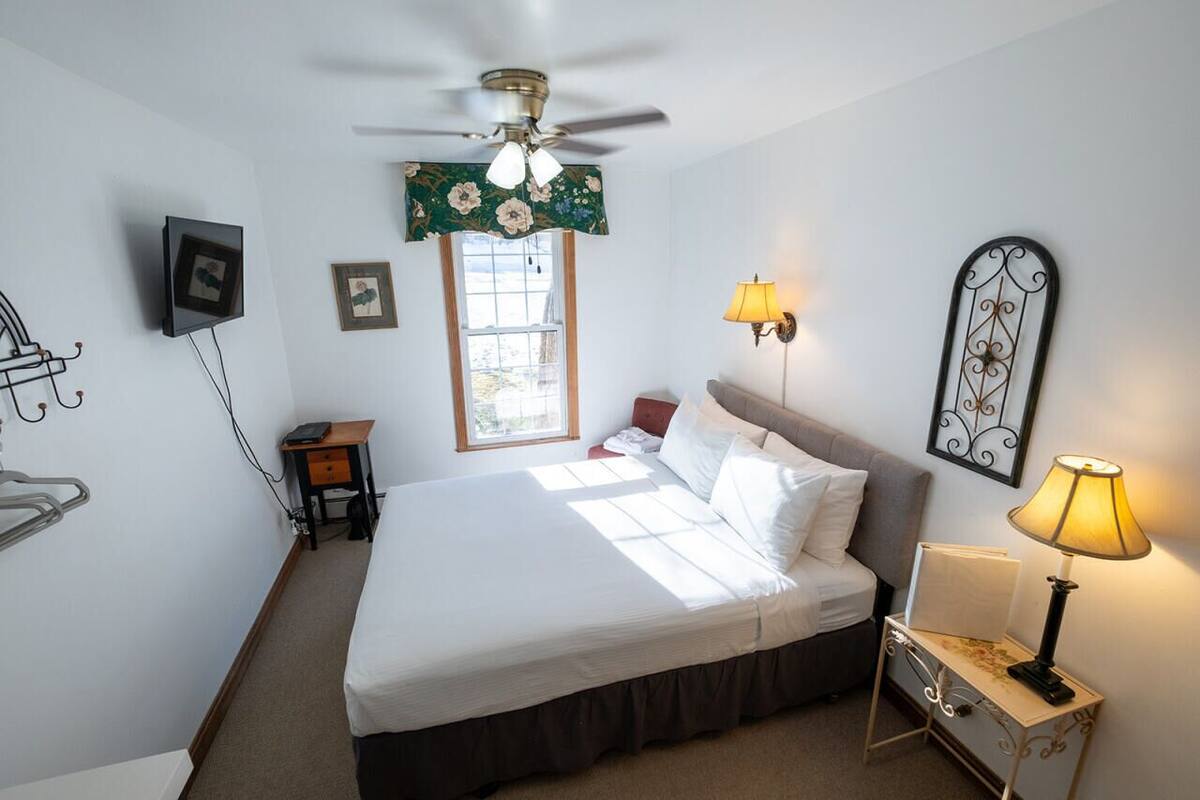 Quarto de hotel com cama, ventoinha no teto, televisão fixada na parede, abajur e janela com luz natural.