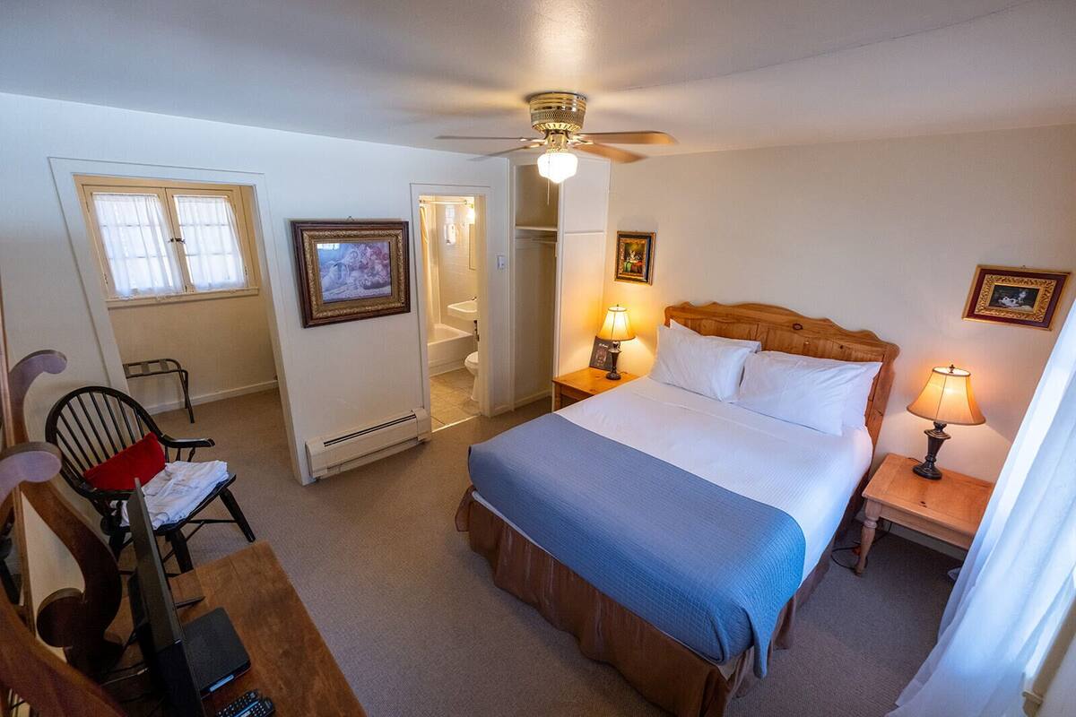 Quarto com uma cama, duas luminárias de cabeceira, ventilador de teto, uma cadeira, escrivaninha com um laptop e porta para um banheiro.