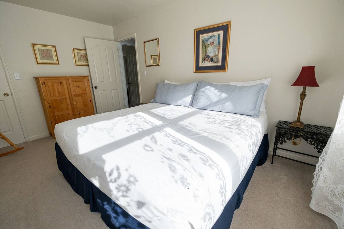 Quarto com cama, guarda-roupa de madeira, porta, quadro emoldurado, luminária e uma pequena mesa.