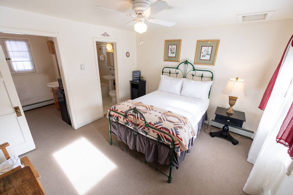 Um quarto com cama, mesa de cabeceira, luminária, ventilador de teto e porta que dá para um banheiro.