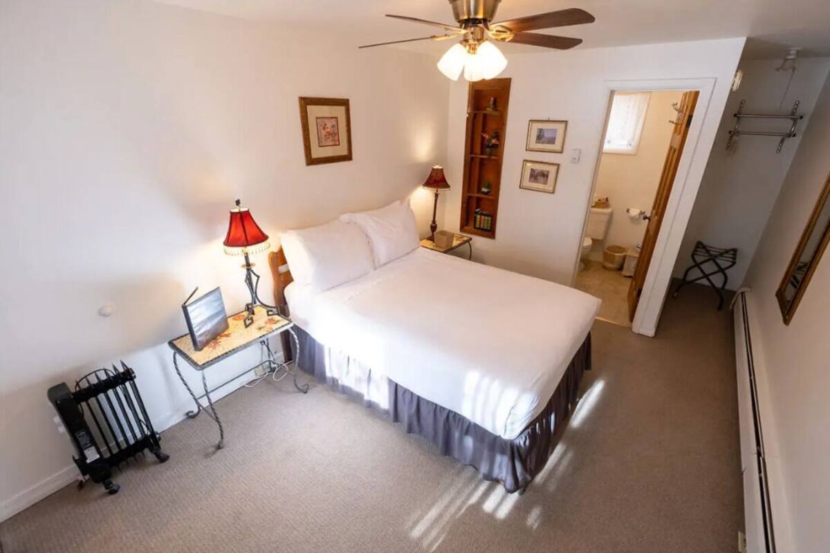 Quarto com cama, mesas de cabeceira, escrivaninha com luminária, ventoinha no teto e radiador.