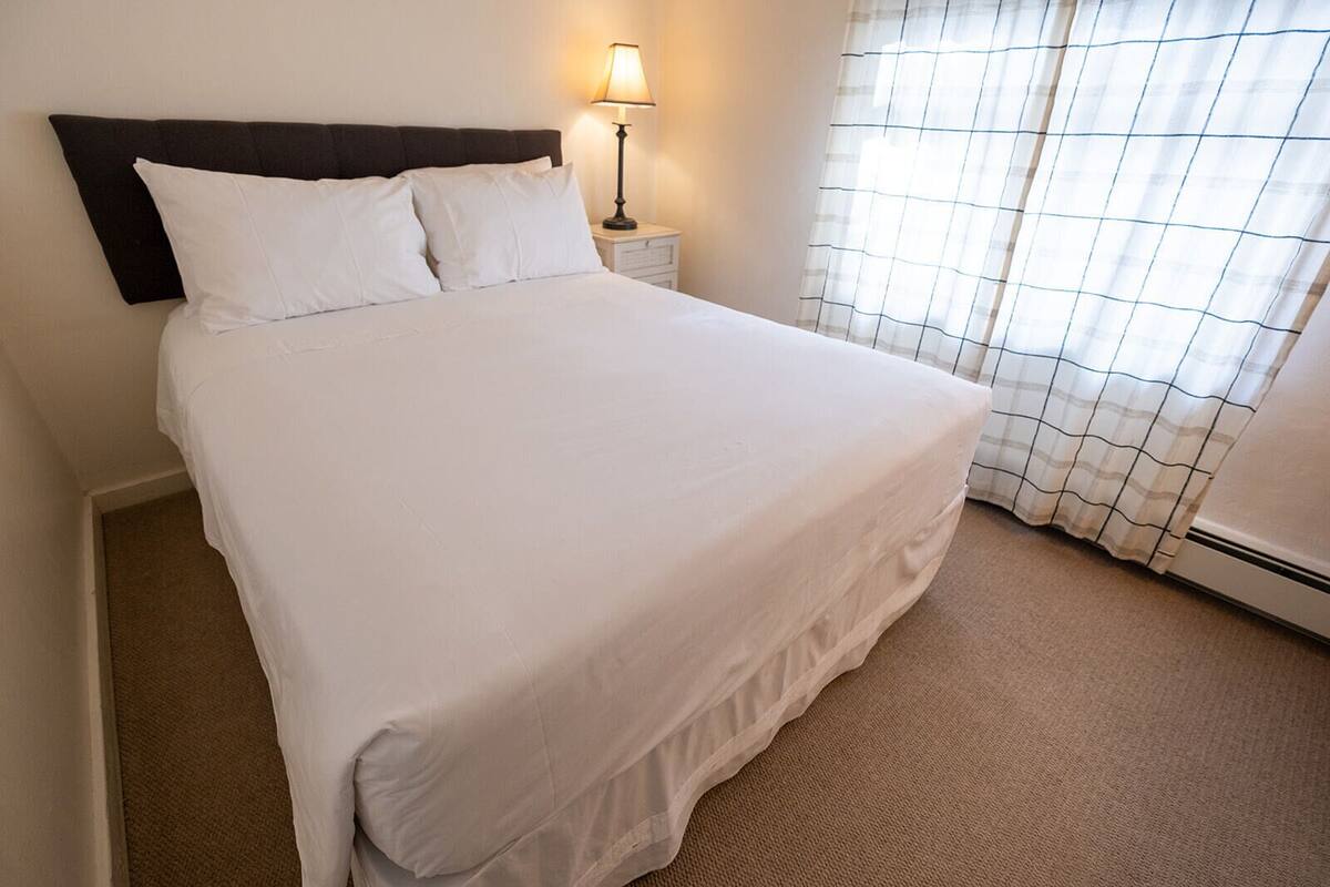 Uma cama bem arrumada com lençóis brancos, cabeceira escura, luminária de cabeceira e janela com cortina xadrez.