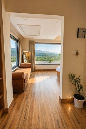 Quarto com piso de madeira, sofá marrom, cama, janela grande com vista para o verde e um vaso de planta.
