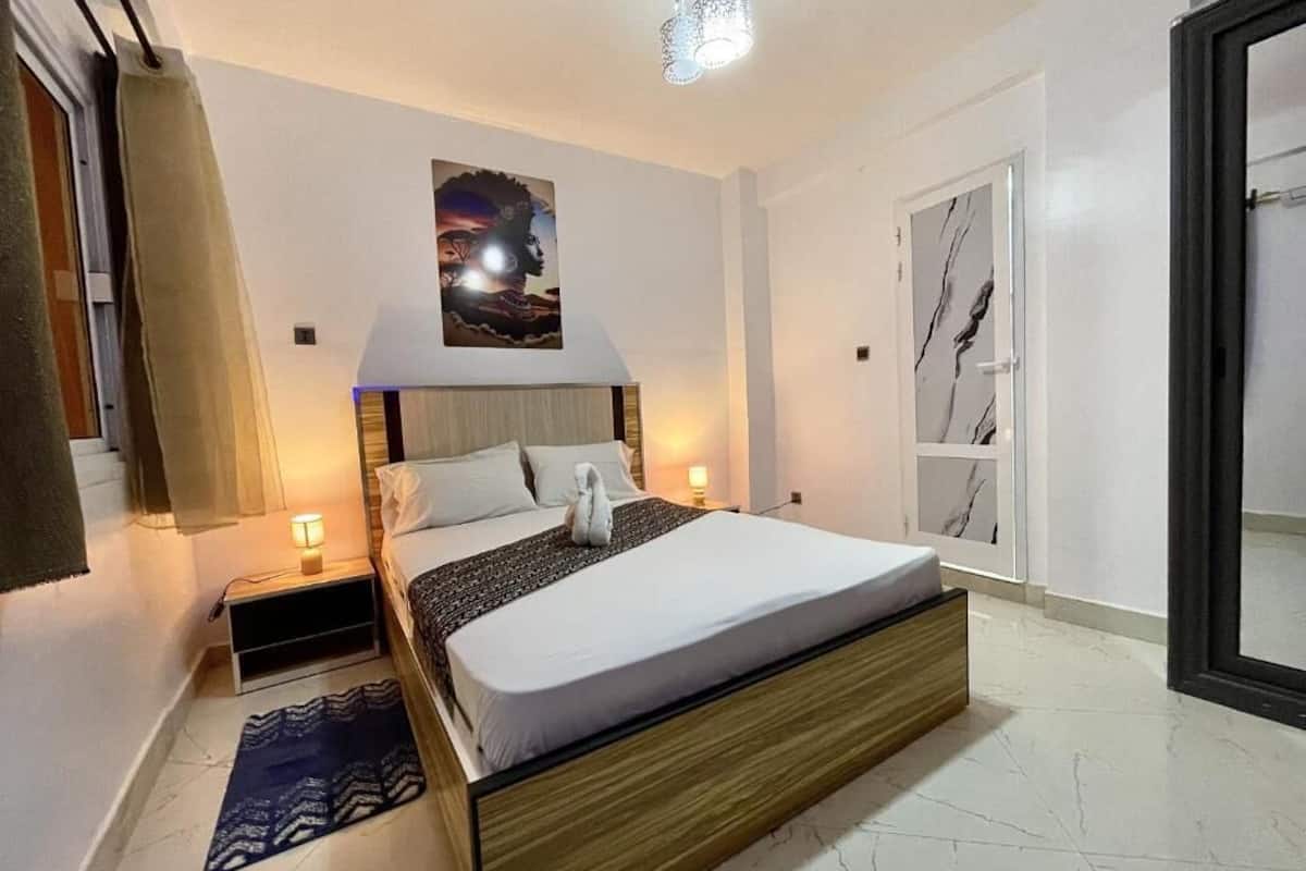 Quarto com cama, luminárias de cabeceira, mesa de cabeceira, obra de arte na parede, porta com painel de vidro e tapete estampado no chão.