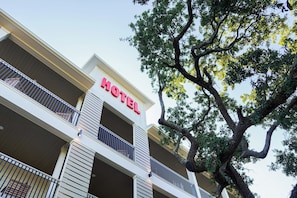 Um hotel com uma placa grande que diz 'HOTEL', várias varandas e uma árvore imponente em primeiro plano.