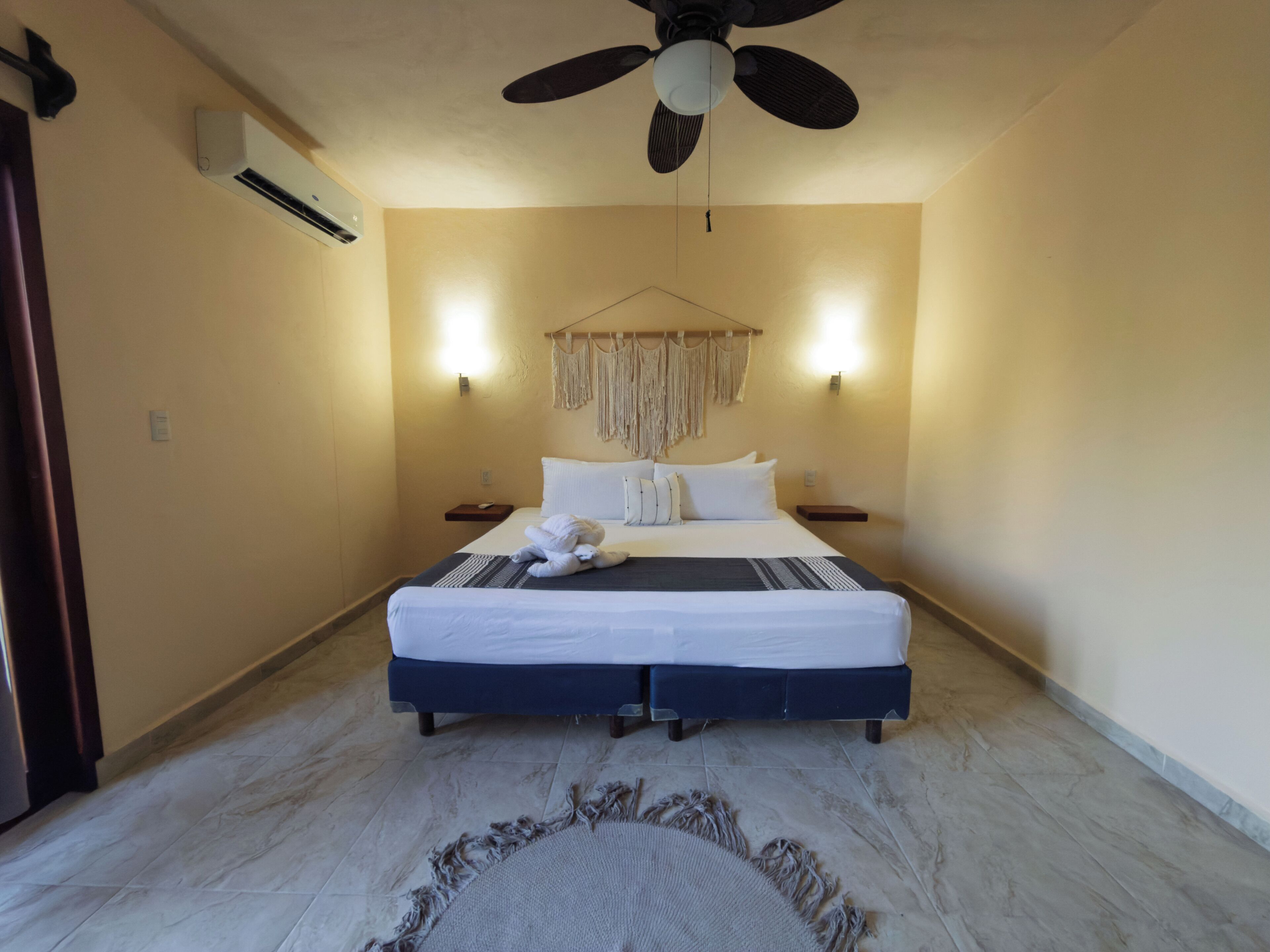 Quarto de hotel com cama, ventoinha no teto, aparelho de ar condicionado, luminária fixada na parede e um pendurar de macramé decorativo.