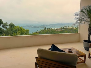 Uma varanda com vista para a paisagem urbana e uma ponte, uma cadeira de madeira, uma pequena mesa com uma tigela e uma planta em vaso.