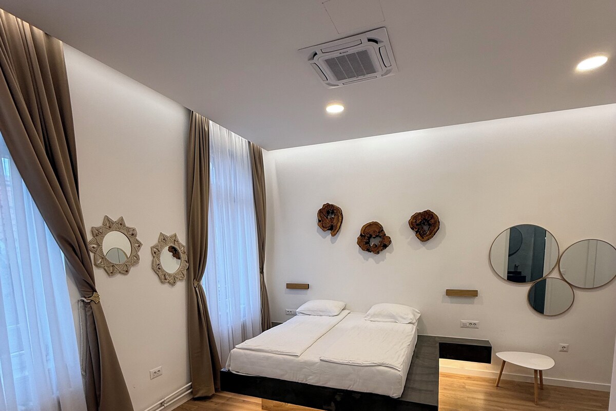 Quarto moderno com cama, mesa de cabeceira pequena, espelho e luminária no teto.