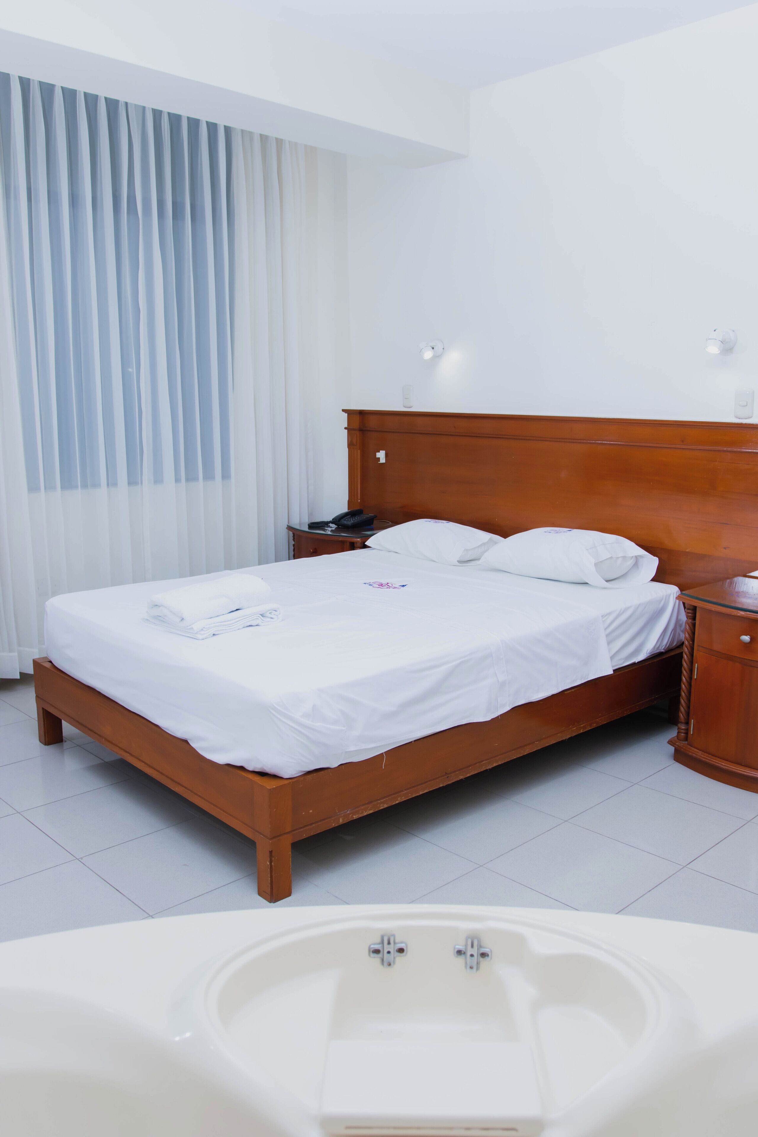 Quarto de hotel com cama de madeira, roupa de cama branca, mesa de cabeceira com telefone e uma banheira grande e branca.