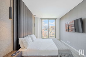 Quarto de hotel moderno com uma cama grande, televisão na parede e vista para edifícios pela janela.