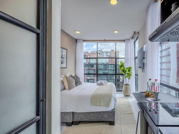 Quarto moderno com cama, planta em vaso e vista da cidade através de grandes janelas.