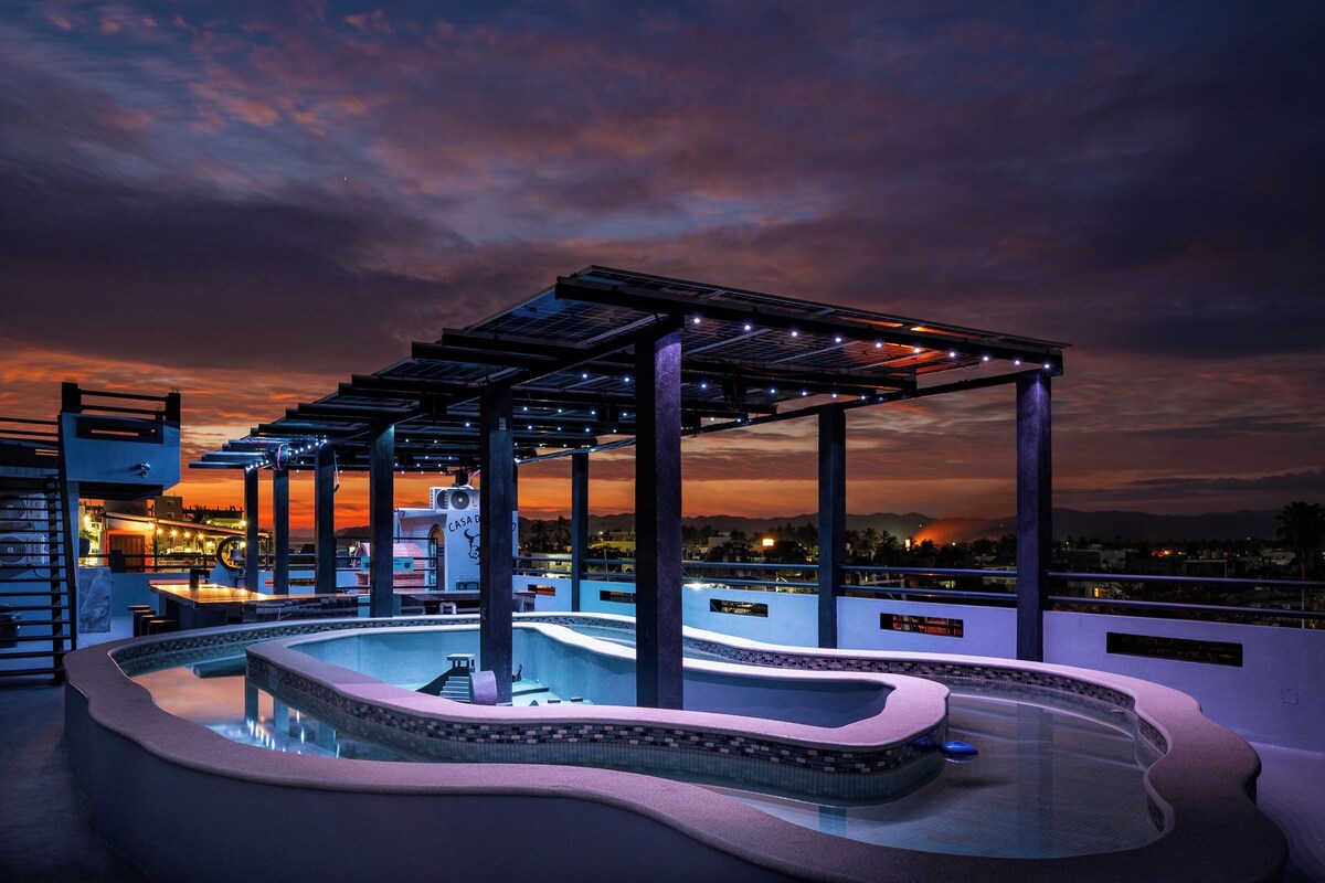 Piscina no terraço com pérgola, banheira de hidromassagem e vista para a cidade ao entardecer.
