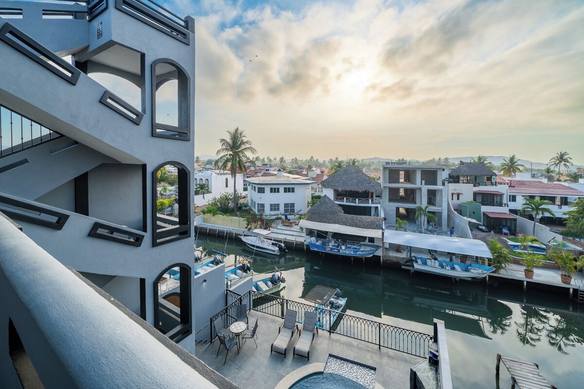 Um edifício moderno com área de piscina, barcos e vista para uma cidade costeira.