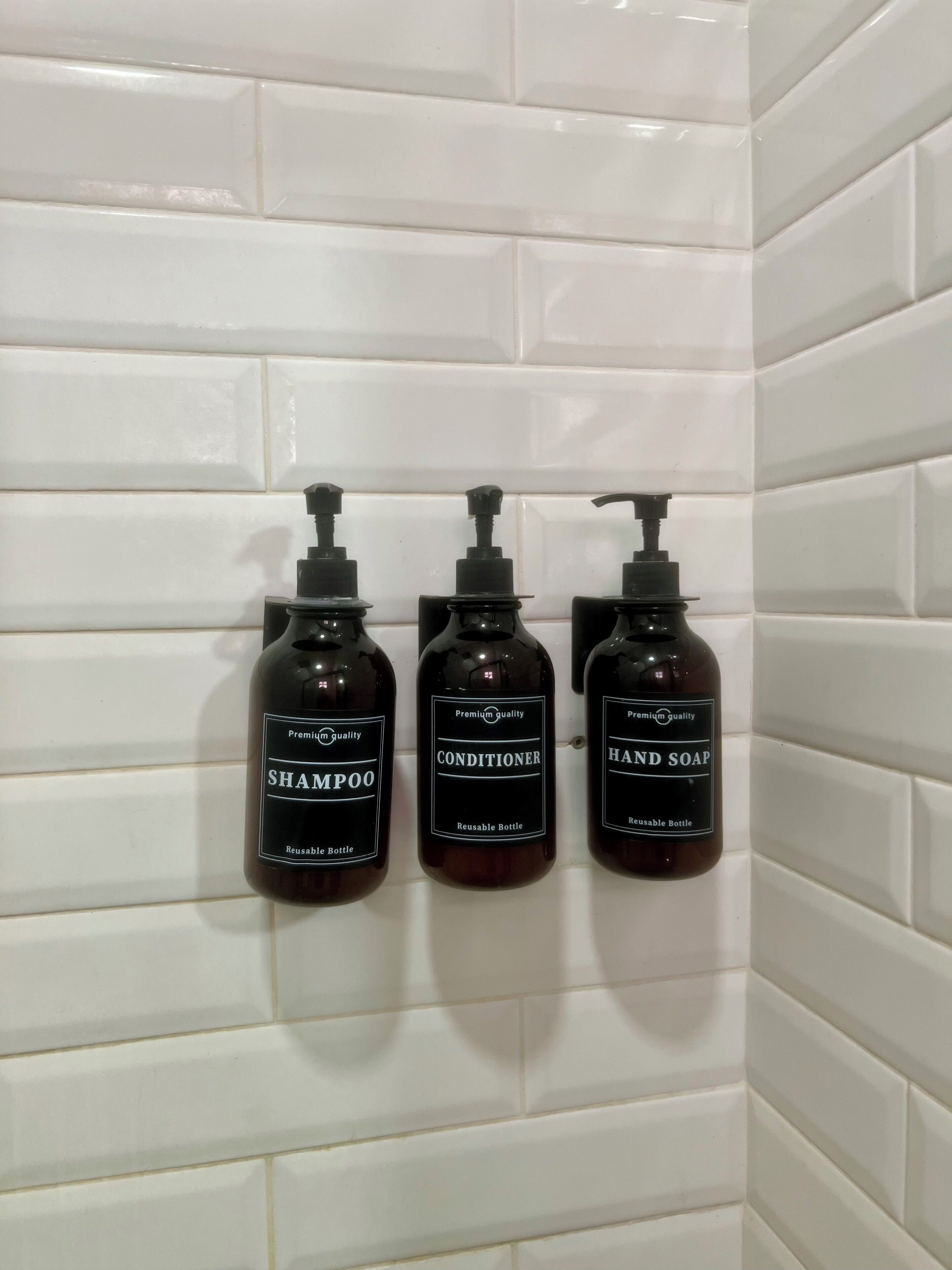 Três garrafas reutilizáveis de shampoo, condicionador e sabonete líquido fixadas numa parede revestida de azulejos.