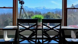 Quarto com um telescópio em um tripé, uma mesa com cadeiras e vista para montanhas cobertas de neve através de grandes janelas.