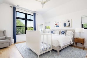 Quarto com berço, cama, mesa de cabeceira, cadeira e uma janela grande com cortinas.