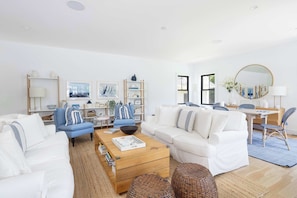 Uma sala de estar moderna com sofás brancos, uma mesa de centro de madeira e um espaço de jantar com uma mesa redonda e cadeiras.