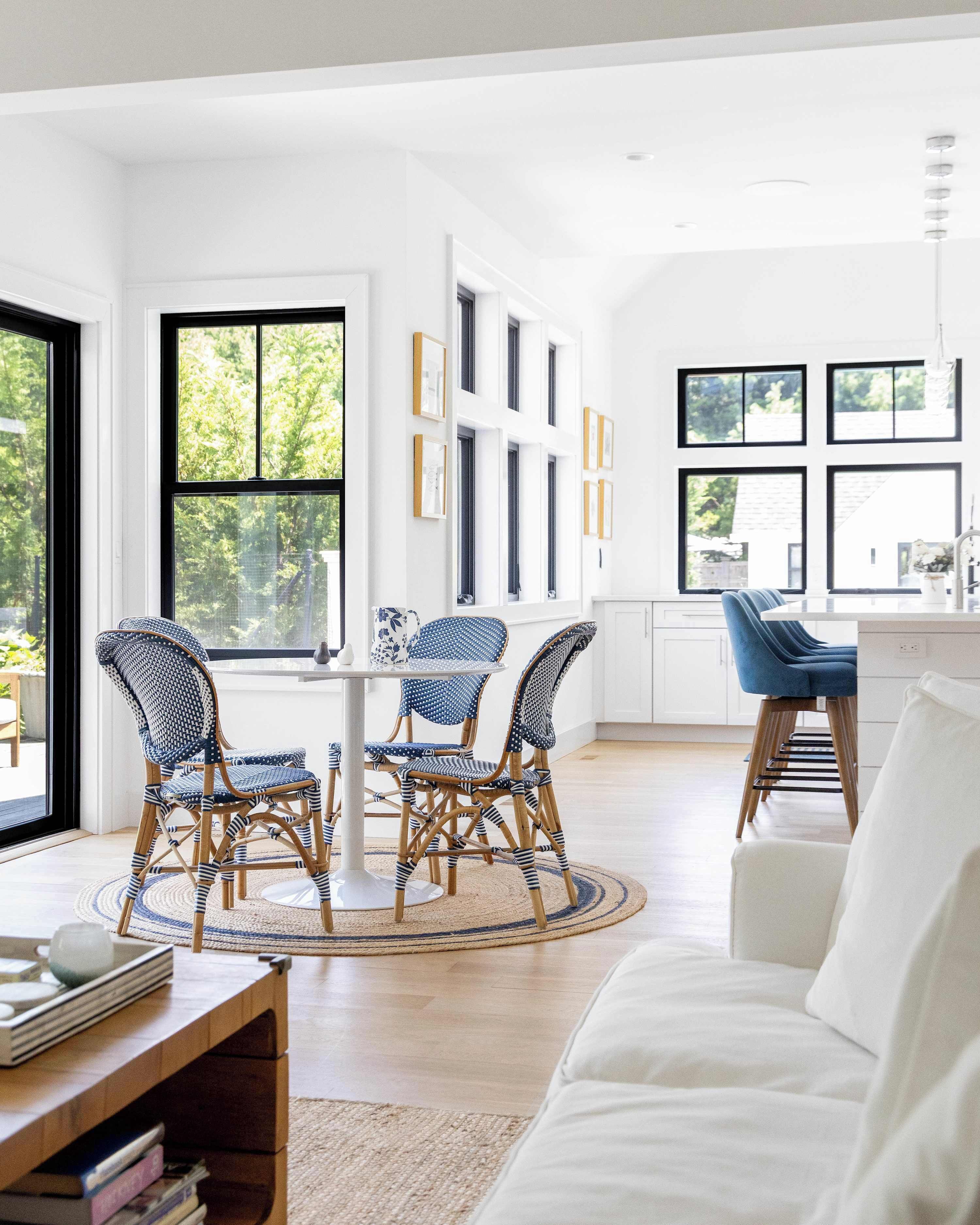 Uma sala de estar moderna com um sofá branco, um espaço de refeições com cadeiras de vime e uma mesa branca, e uma cozinha com armários brancos e um banco de bar azul.