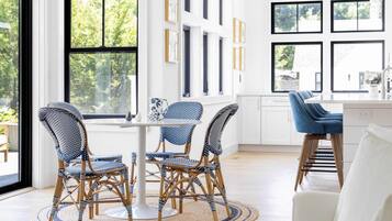 Uma sala de estar moderna com um sofá branco, um espaço de refeições com cadeiras de vime e uma mesa branca, e uma cozinha com armários brancos e um banco de bar azul.