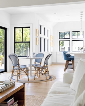 Uma sala de estar moderna com um sofá branco, um espaço de refeições com cadeiras de vime e uma mesa branca, e uma cozinha com armários brancos e um banco de bar azul.