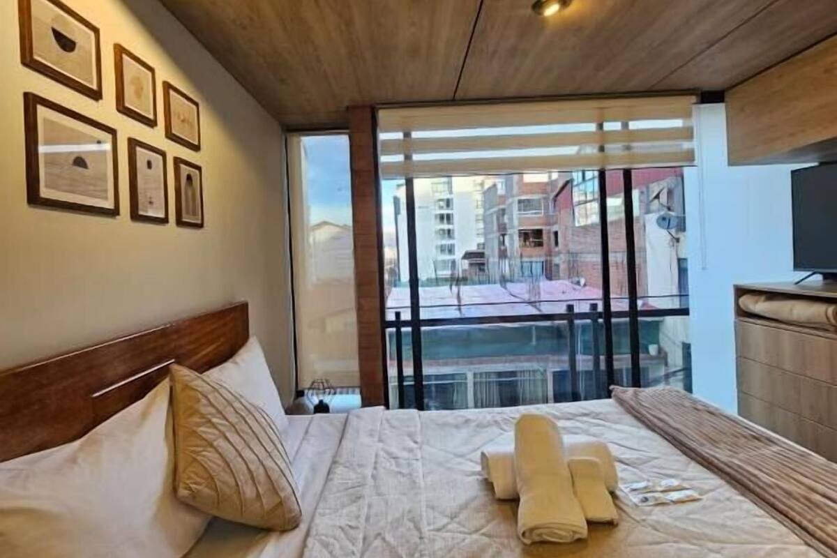 Quarto de hotel com teto de madeira, uma cama grande com cabeceira, mesa de cabeceira, televisão e varanda com vista para edifícios.