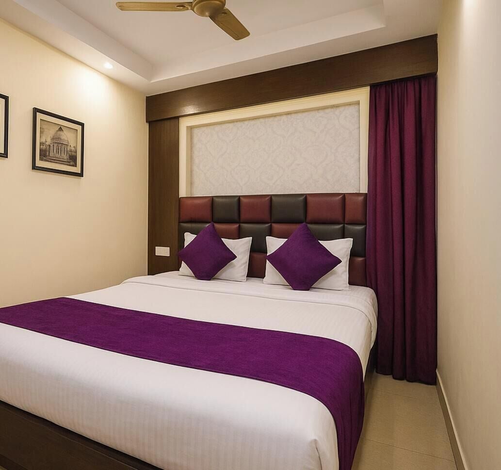 Quarto de hotel com cama, ventoinha no teto, janela com cortinas e um quadro pendurado na parede.
