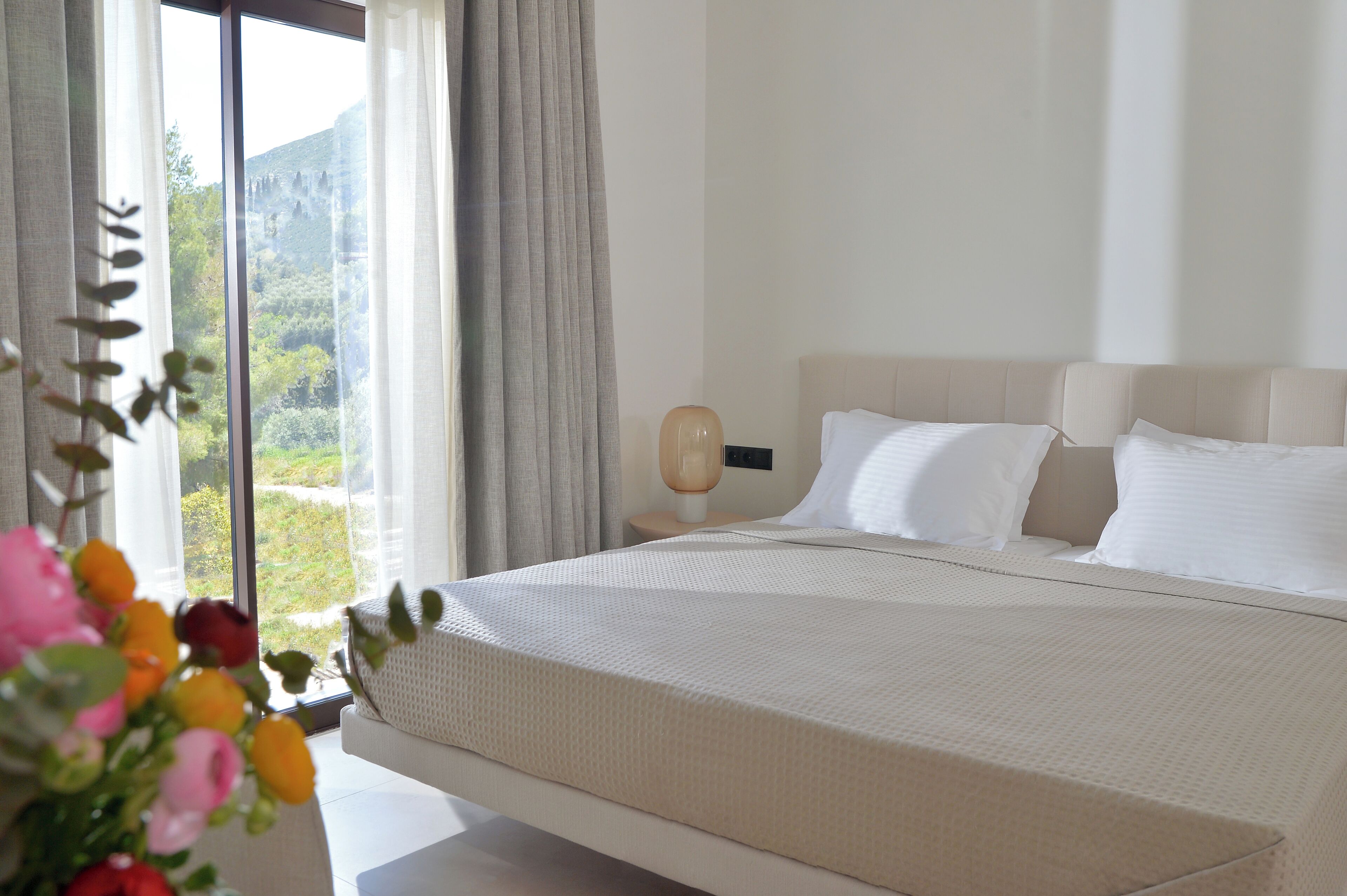 Quarto com uma cama grande, mesa de cabeceira com abajur e vista para o verde pela janela.