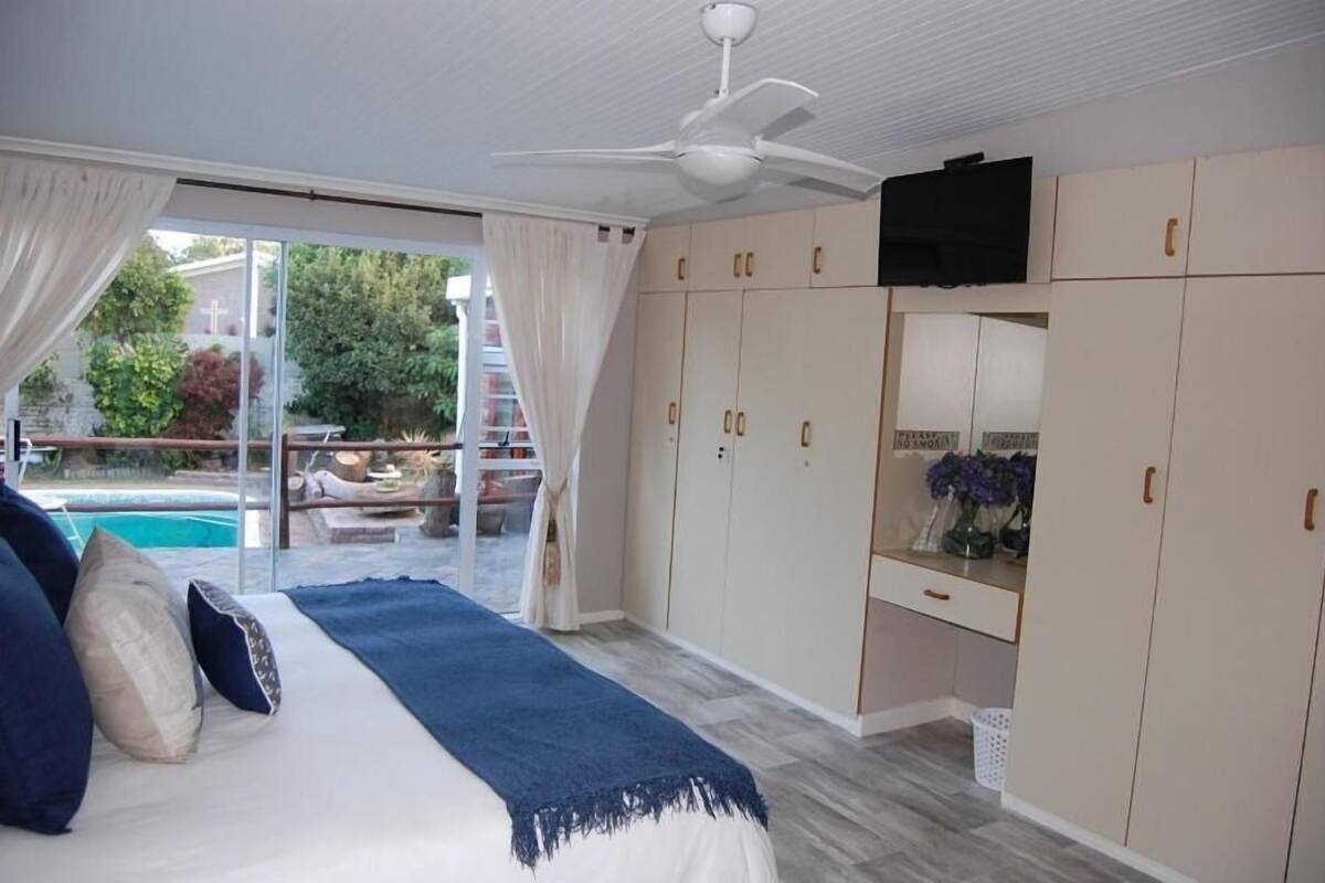 Quarto com cama, ventilador de teto, TV sobre um móvel e vista para uma piscina.