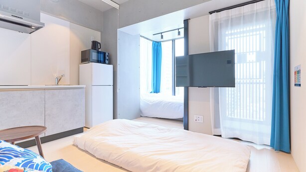 Quarto de hotel moderno com cama, sofá, televisão, cozinha compacta e janela com cortinas.