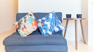 Um sofá cinzento com duas almofadas estampadas e uma pequena mesa de madeira com duas chávenas azuis.