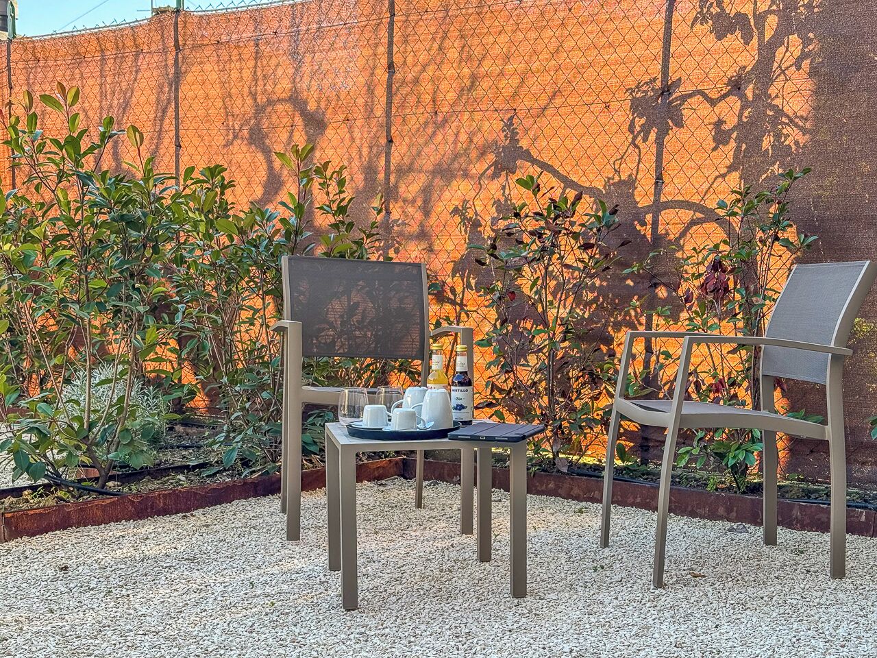 Uma área de pátio com duas cadeiras e uma mesa disposta com uma garrafa, copos e xícaras.