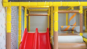 Uma área de recreação infantil com um escorrega vermelho, paredes com rede e estruturas acolchoadas.