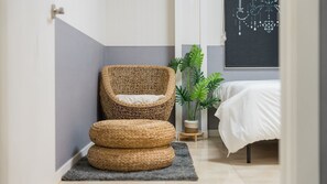 Uma cadeira de vime com almofada, uma planta em vaso e uma cama com roupa de cama branca.