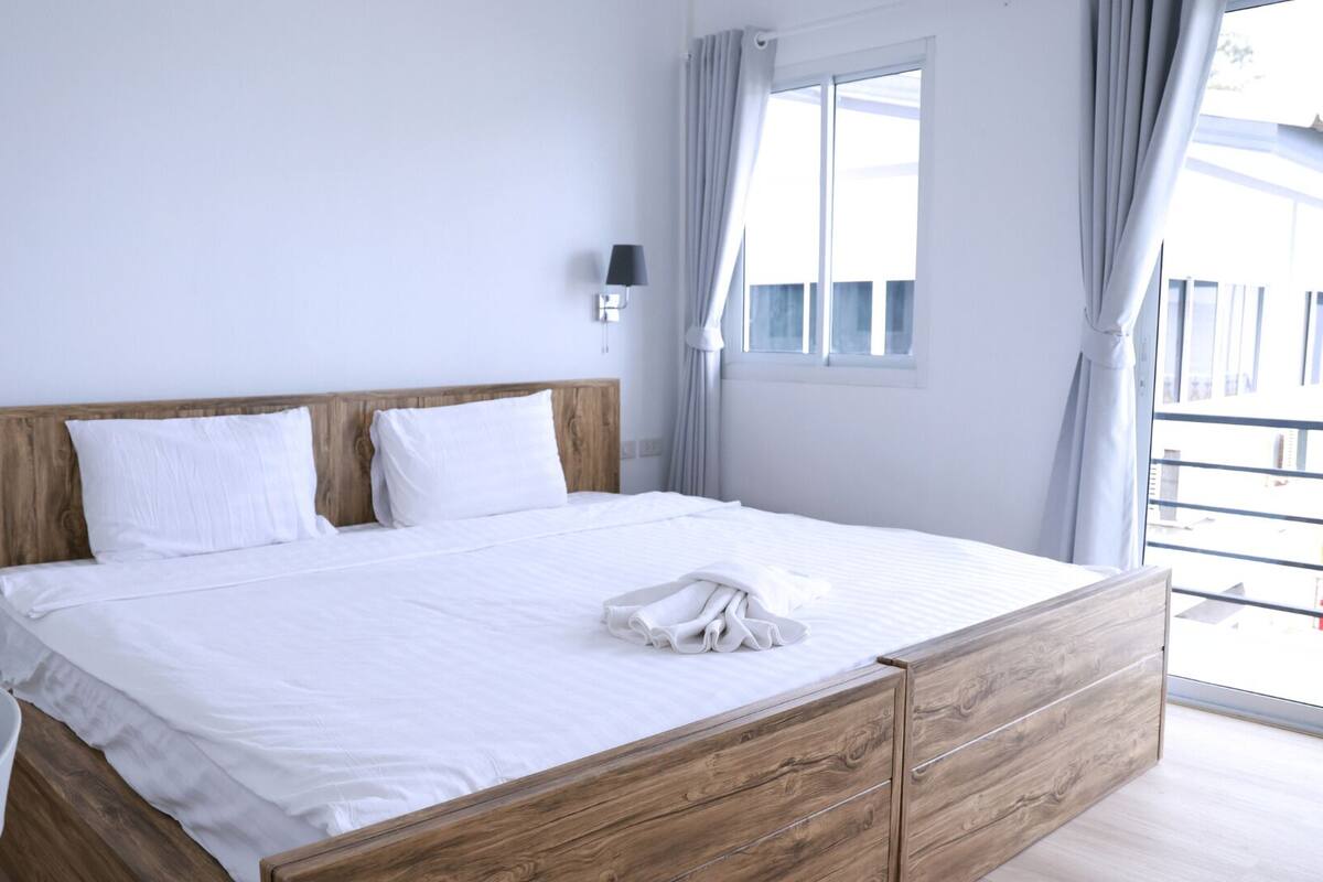 Quarto com estrutura de cama de madeira, roupa de cama branca, mesa de cabeceira, abajur e janela com cortinas.