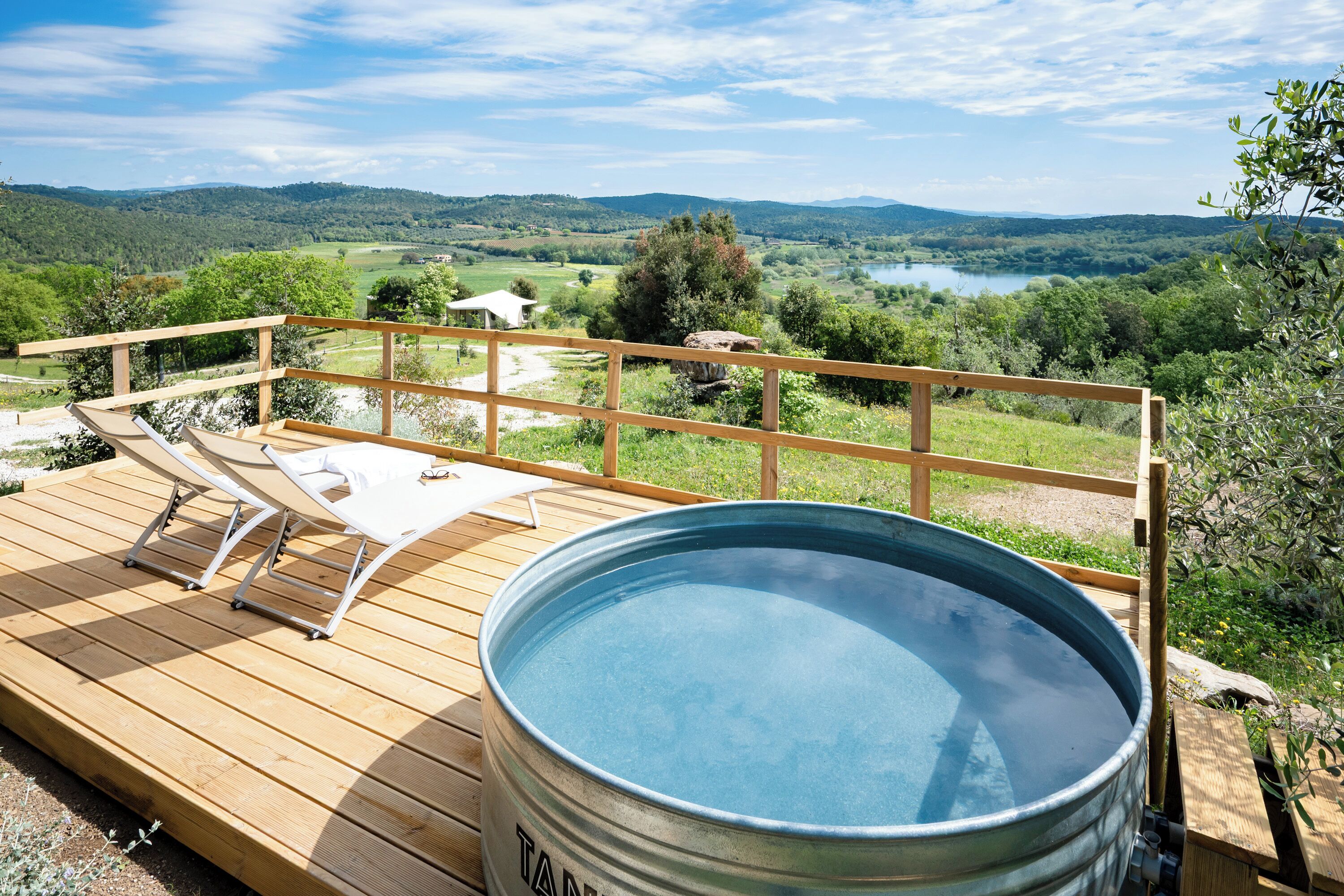 Um deck de madeira com uma banheira de hidromassagem, uma espreguiçadeira e vista para um lago e colinas.