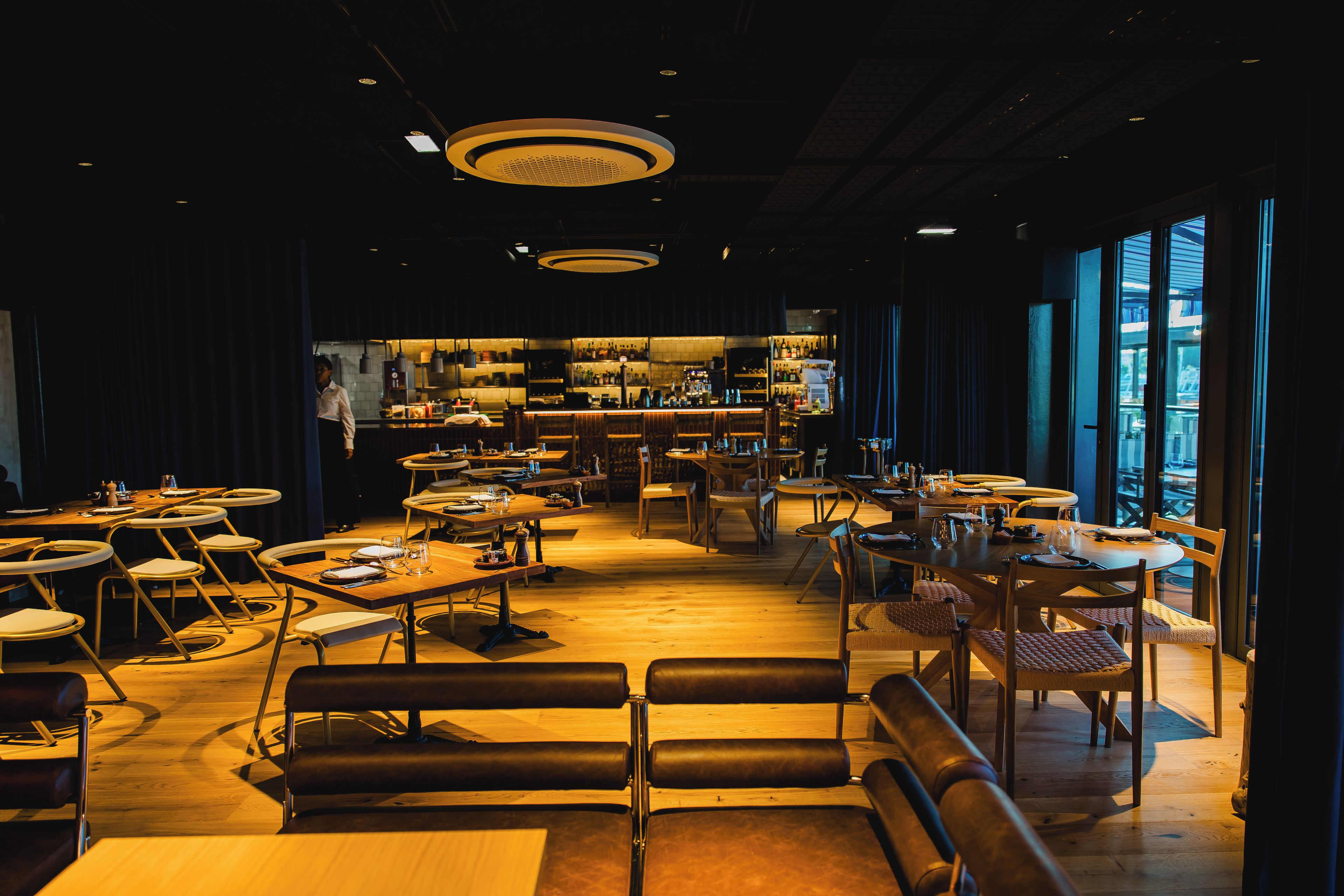Interior de restaurante com várias mesas e cabines de jantar, área de bar e vista através de grandes janelas.