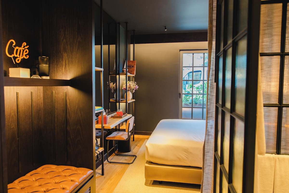 Quarto de hotel com uma cama, uma mesa com uma cadeira, uma pequena mesa com um vaso de flores e uma janela com vista para o exterior.