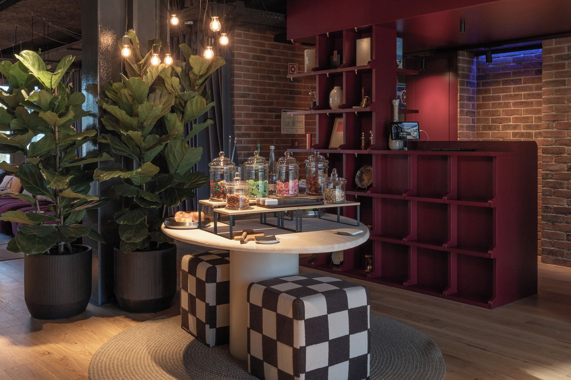 Um interior moderno com uma mesa redonda, pufes xadrez, uma parede de tijolos e uma estante vermelha.
