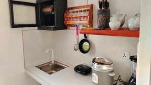 Uma cozinha compacta com pia, cozidela de arroz, liquidificador e uma prateleira com utensílios de cozinha.