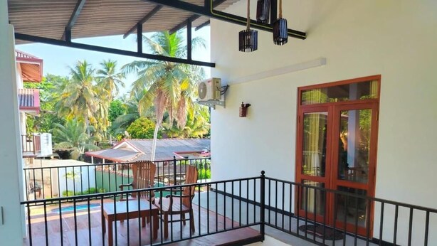 Uma varanda com mesa e cadeiras, uma porta vermelha e vista para palmeiras e um edifício.