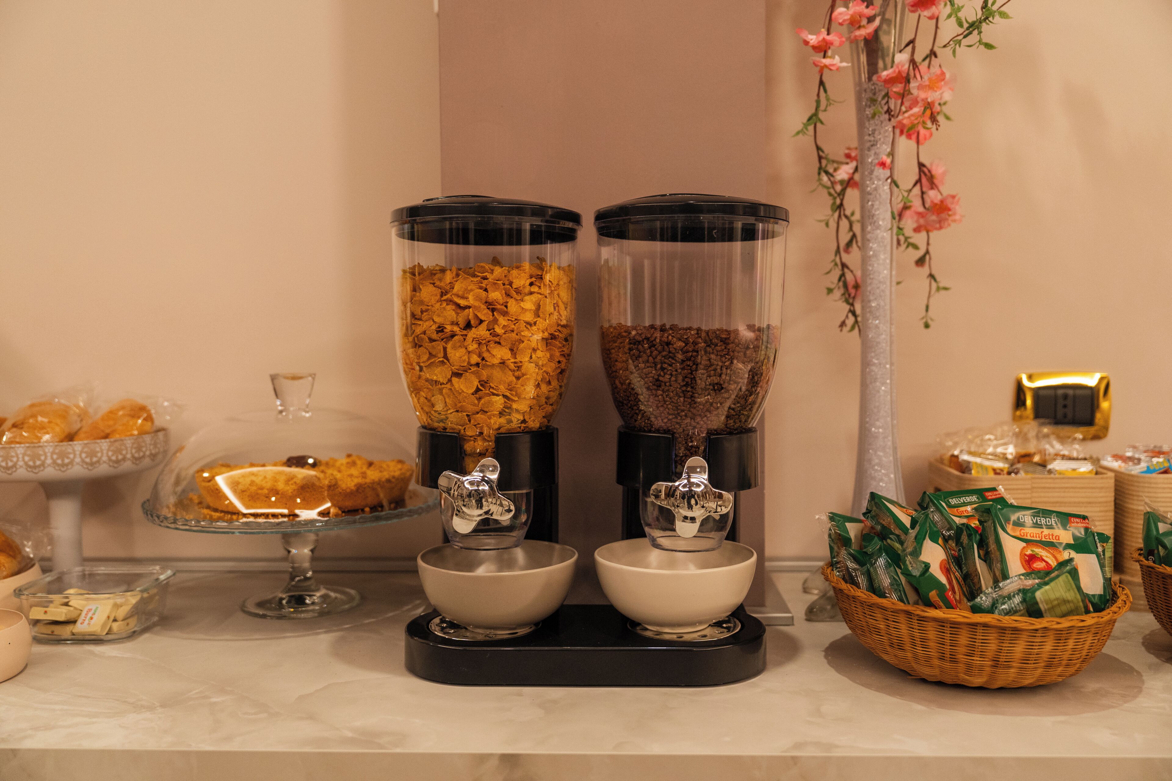 Disposição de buffet com dispensadores de cereais e granola, uma tigela de vidro e uma cesta de produtos embalados.