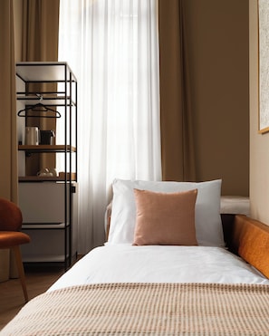 Quarto de hotel bem arrumado, com cama, mesa de cabeceira, prateleira suspensa e uma cadeira.