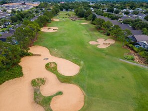 Vista aérea de um campo de golfe, com bunkers de areia, green de aproximação e áreas residenciais ao redor.