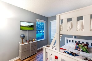 Quarto de criança com cama beliche, televisão, janela com persianas, cômoda e escrivaninha com brinquedos.