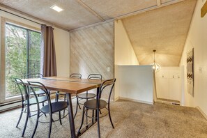 Um quarto com uma mesa e cadeiras de madeira, uma janela grande com cortinas, teto inclinado e luminária pendente.