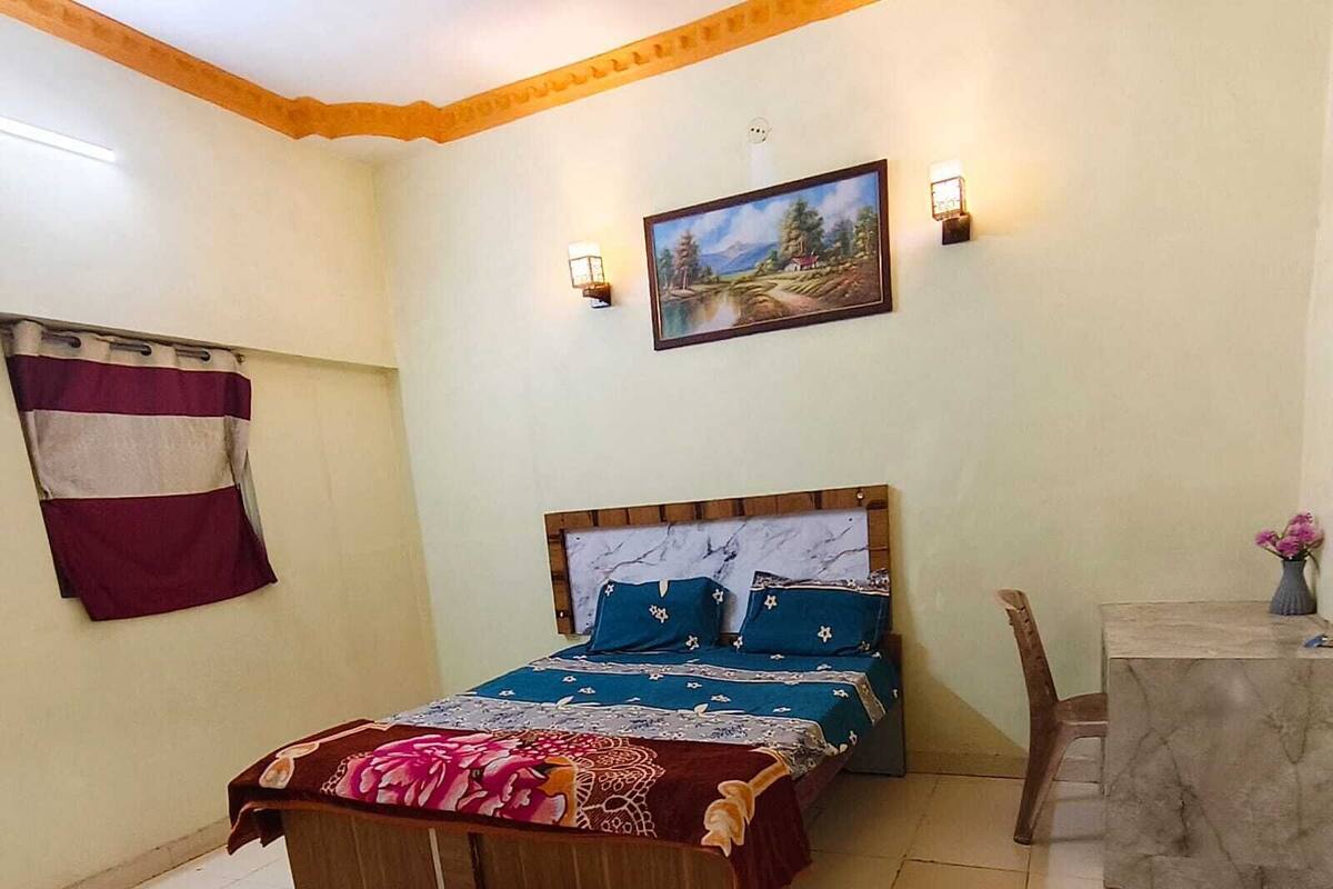 Quarto com cama, uma mesinha, uma cadeira, um quadro na parede e uma cortina.